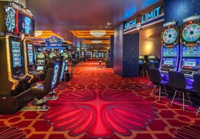 Um salão de jogos de cassino com máquinas caça-níqueis, mesas de blackjack e um corredor com carpete vermelho.
