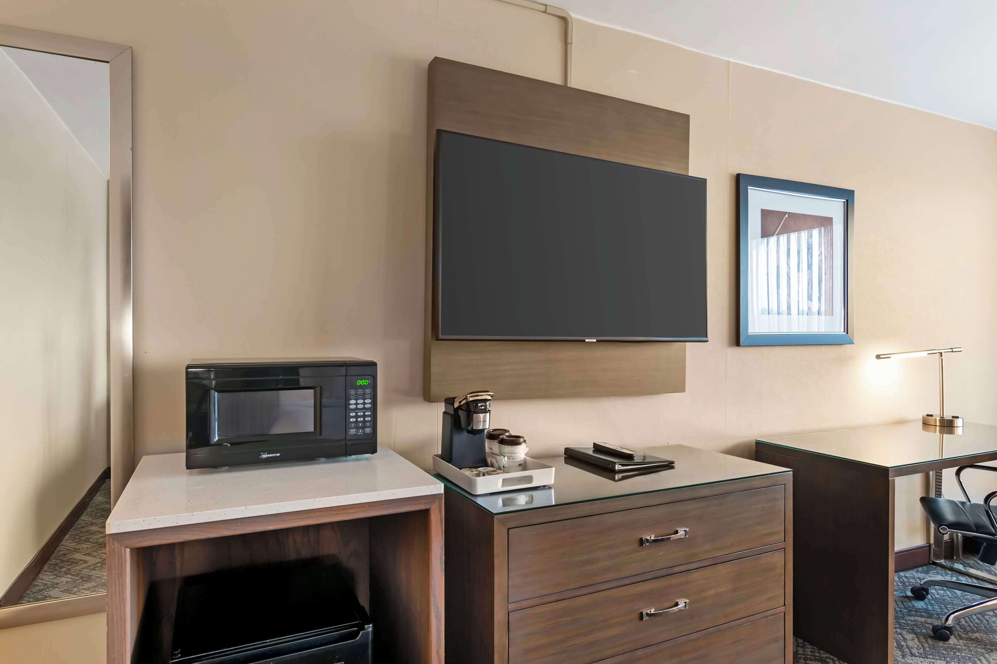Quarto de hotel com cozinha compacta, televisão de tela plana, cafeteira, escrivaninha com cadeira e quadro decorativo na parede.