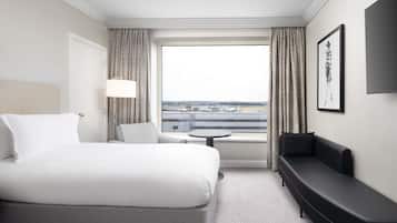 Quarto de hotel com cama, sofá, poltrona, mesa pequena, televisão e janela com vista para um aeroporto.