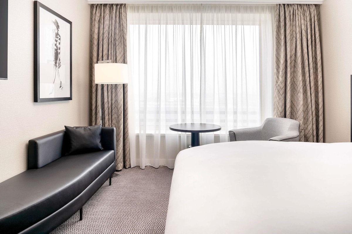 Quarto de hotel com cama, sofá, cadeira, mesa redonda pequena e luminária.
