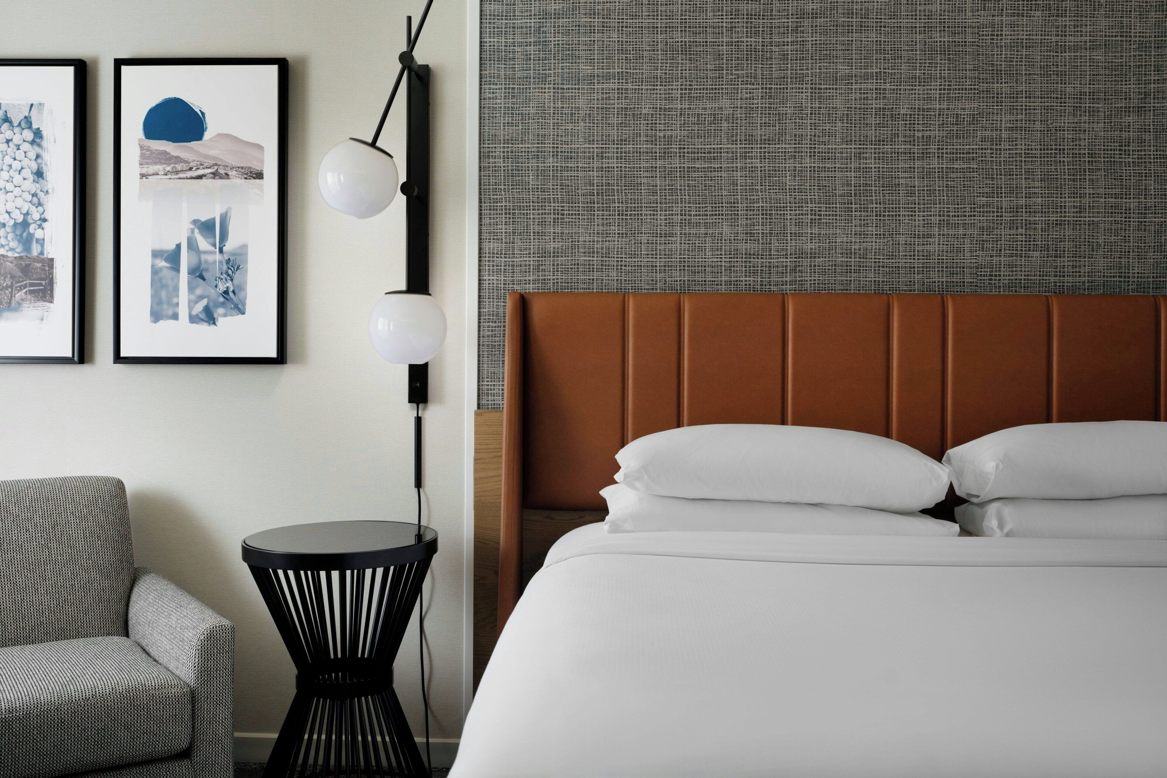 Quarto de hotel com uma cama, uma cadeira, uma mesa redonda e duas molduras na parede.