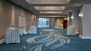 Corredor de hotel com carpete azul e um grande design abstrato. Há mesas com toalhas brancas e velas, e o corredor dá acesso a uma sala identificada como “Sale”.