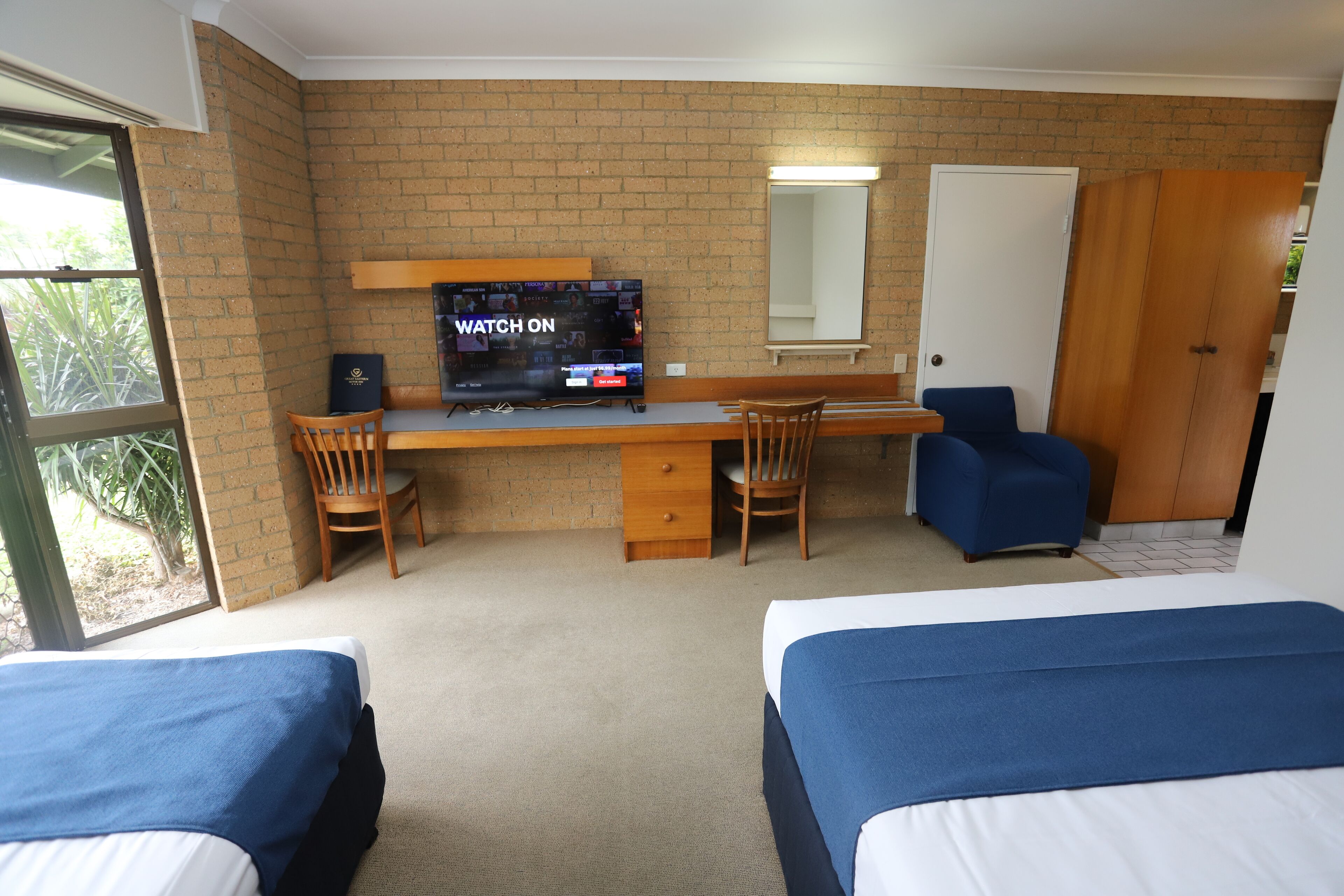 Quarto de hotel com duas camas, uma mesa com televisão, uma cadeira e um guarda-roupa.