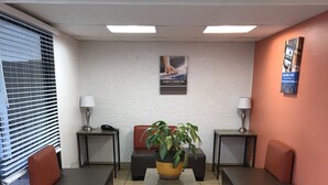 Uma área de espera com piso de azulejos, dois sofás pretos, uma pequena mesa com uma planta em vaso e uma placa fixada na parede.