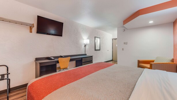 Quarto de hotel com uma cama, uma escrivaninha, uma cadeira, uma televisão fixada na parede e uma porta que dá para outro quarto.