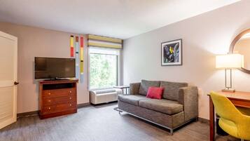 Quarto de hotel moderno com sofá cinzento, cómoda de madeira, televisão de ecrã plano, secretária com candeeiro e janela com vista para vegetação.