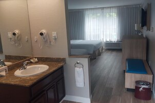 Casa de banho de hotel com lavatório, espelho, porta-toalhas e uma cama visível pela janela.