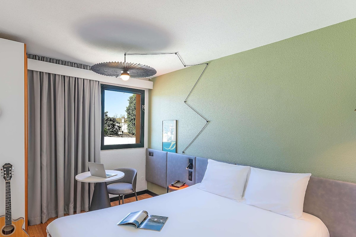 Quarto de hotel com uma cama, uma escrivaninha com um laptop, um violão e vista para o exterior pela janela.