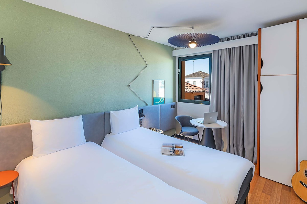 Quarto de hotel com uma cama grande, uma pequena mesa com um laptop, uma cadeira, um violão e uma janela com cortinas.