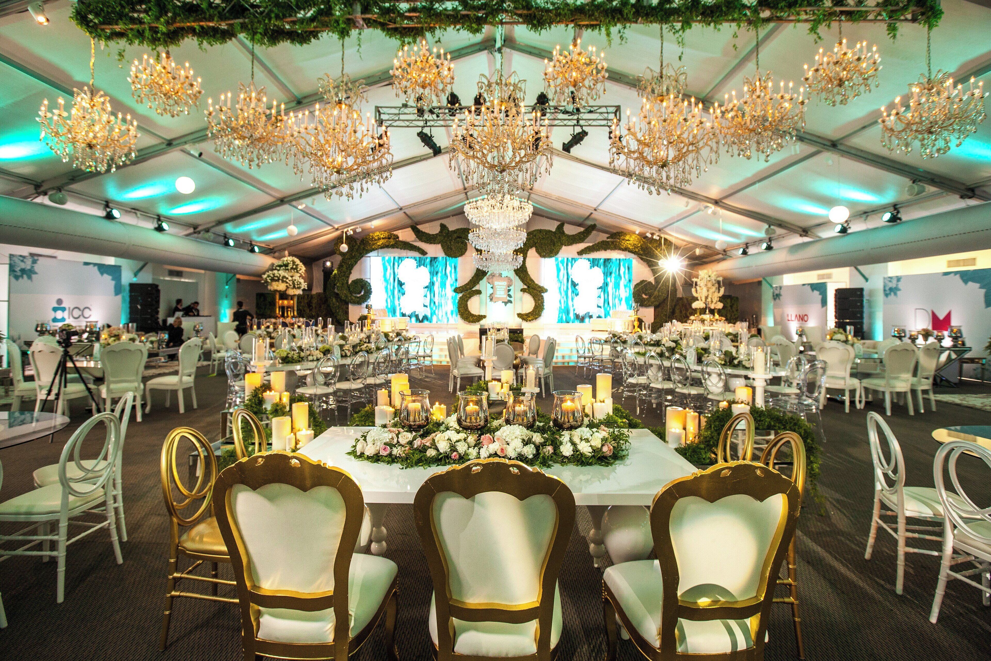 Salão de banquetes com várias mesas montadas para um evento formal, com lustres, centros de mesa florais e cadeiras elegantes.