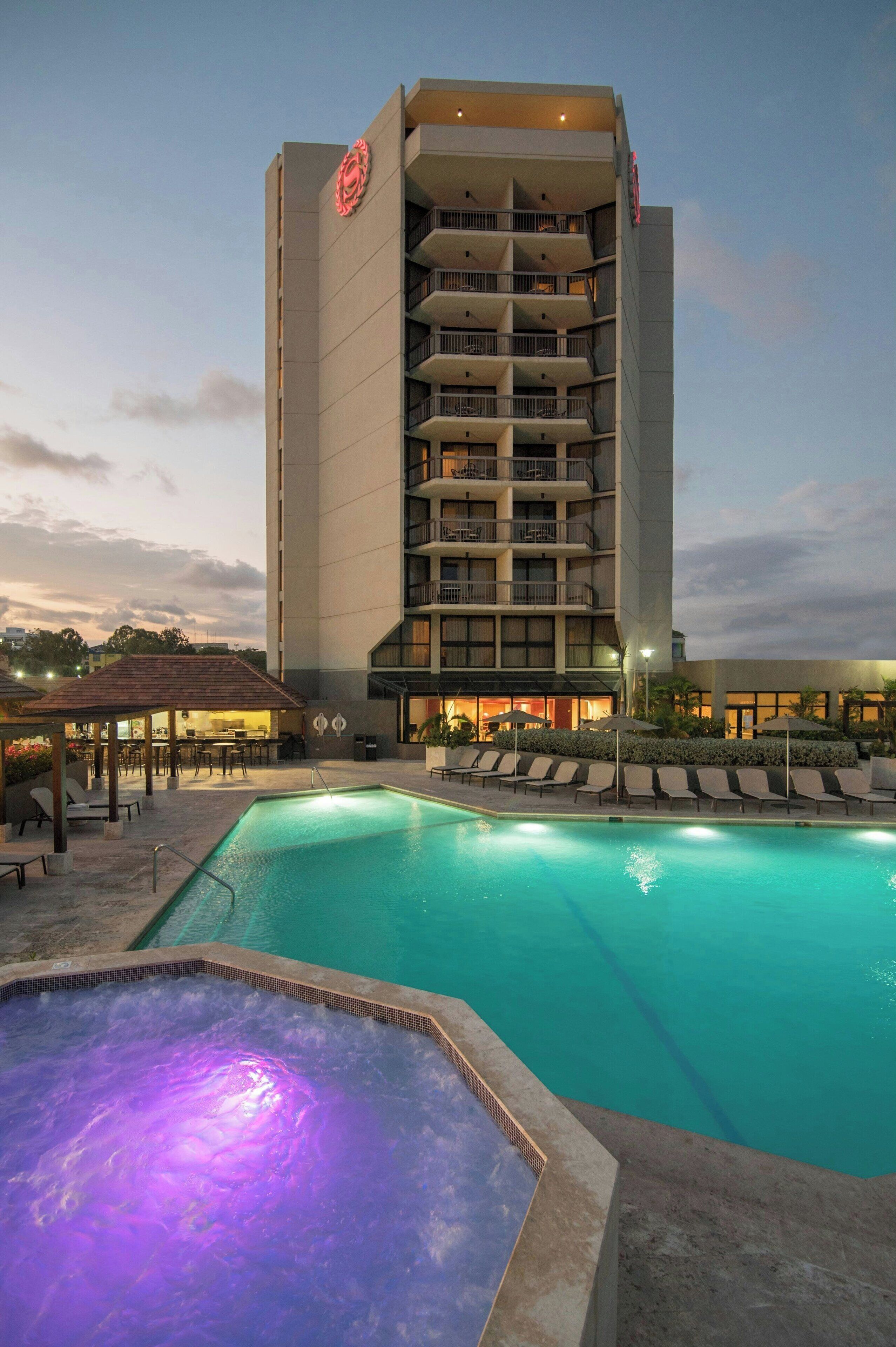 Hotel com uma grande piscina ao ar livre, área de estar coberta e fachada de edifício bem iluminada.