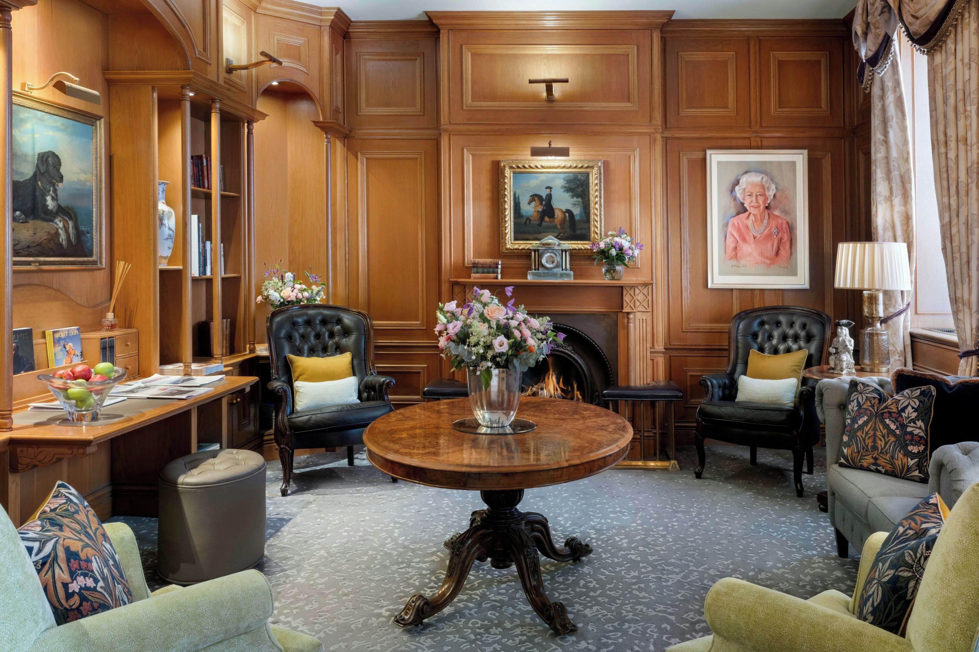 Sala elegante com lareira, dois poltronas, uma mesa redonda e uma estante de livros com livros e um vaso de flores.