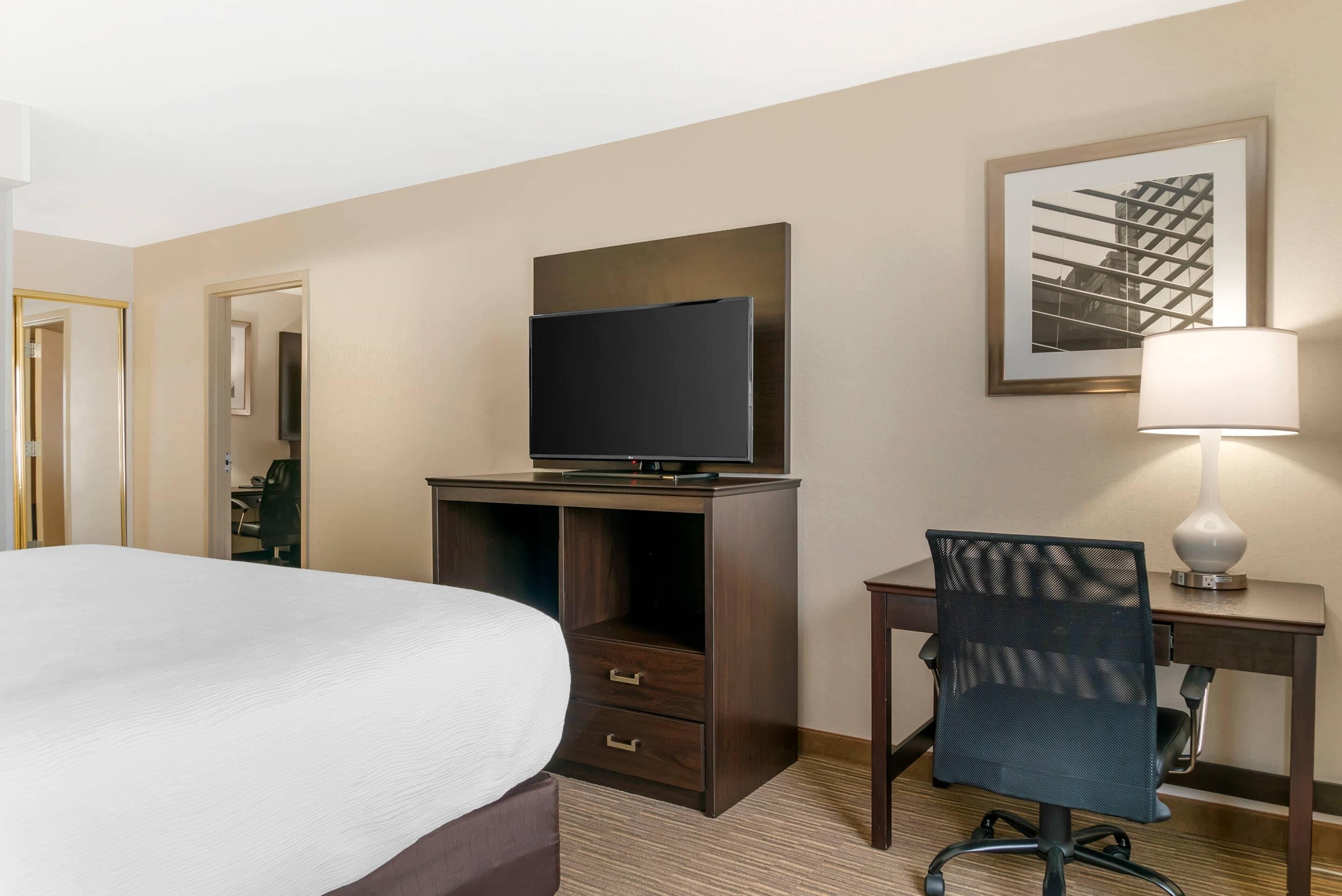 Quarto de hotel com cama, escrivaninha com cadeira, televisão e abajur.