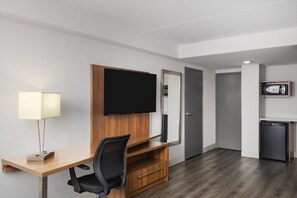 Quarto de hotel com escrivaninha de madeira, uma cadeira, televisão na parede, porta, micro-ondas e um pequeno frigorífico.