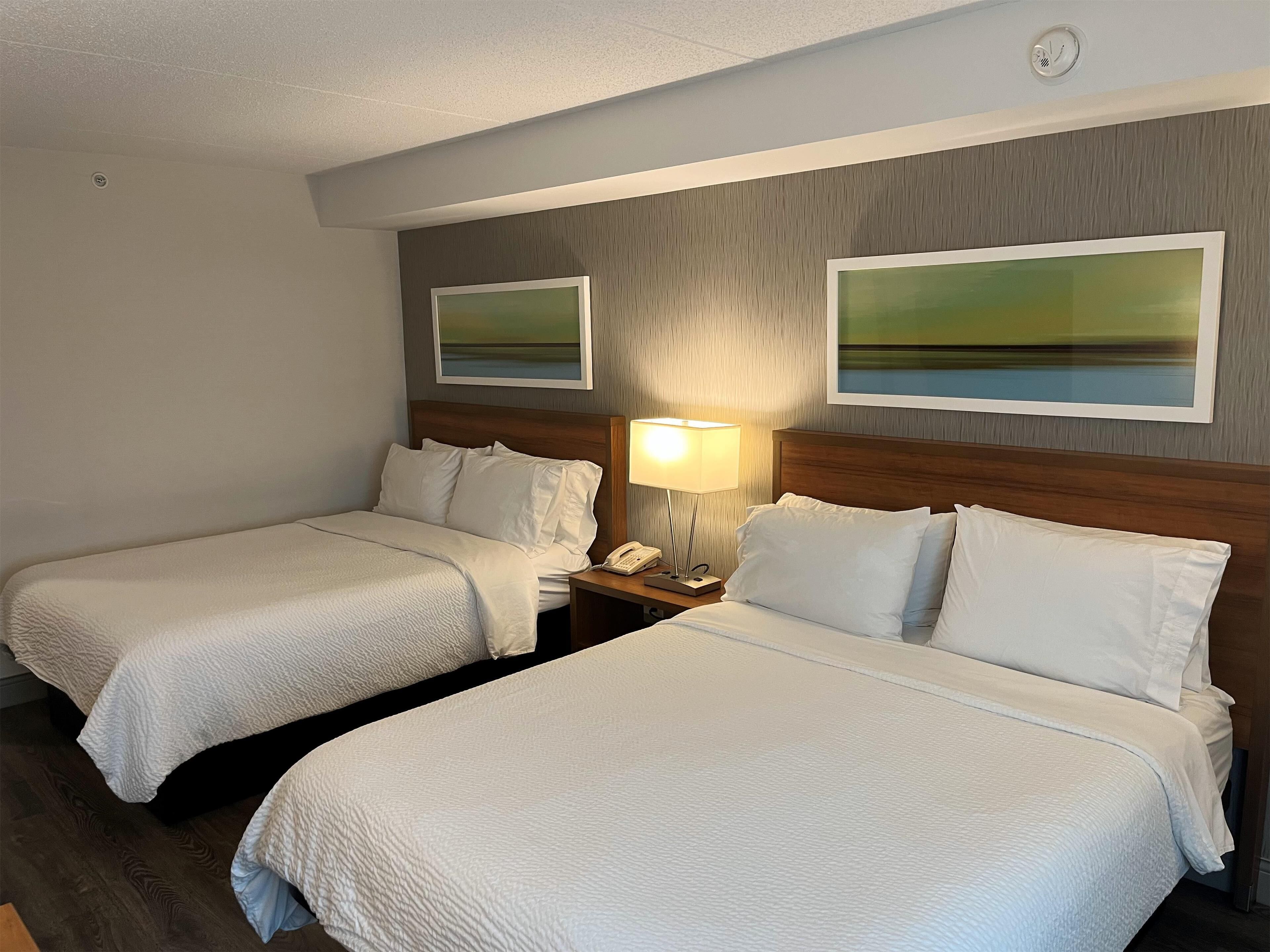 Quarto de hotel com duas camas, uma mesinha de cabeceira com abajur e duas imagens emolduradas na parede.