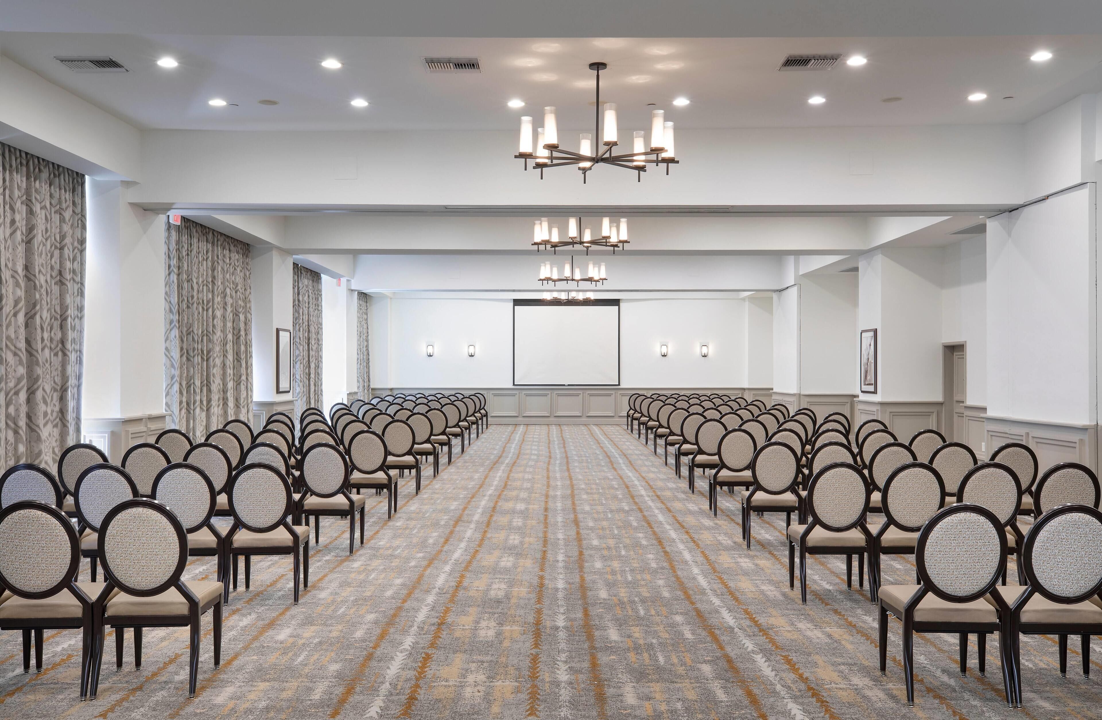 Uma sala de conferências com fileiras de cadeiras, uma tela grande e um lustre.