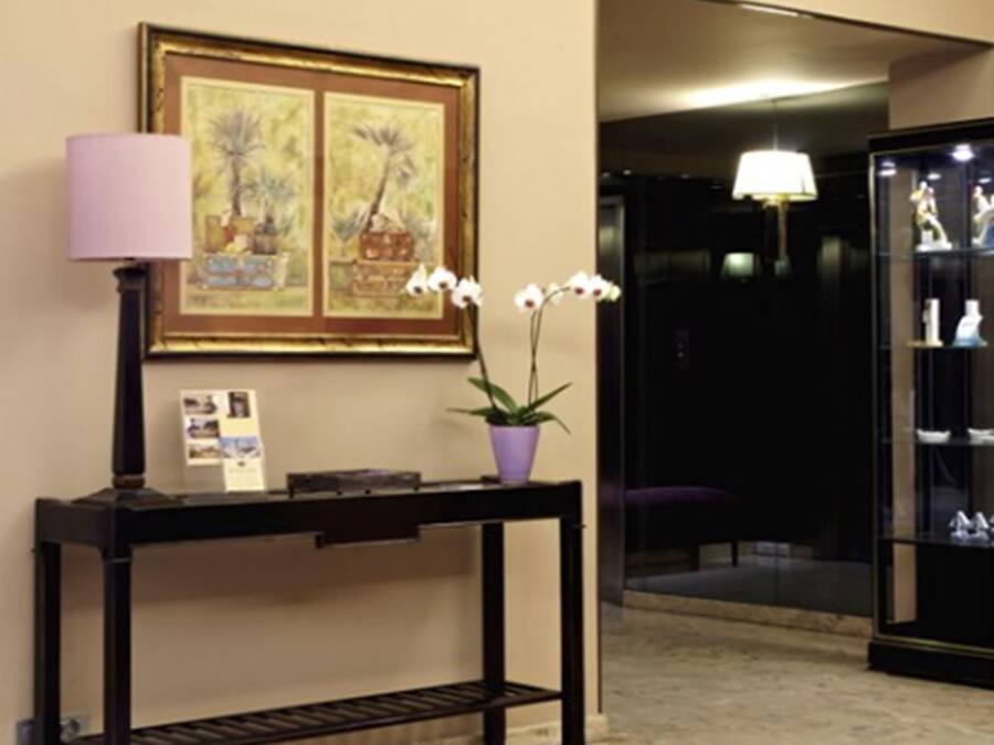 Um hall de entrada com um quadro emoldurado, uma mesa com abajur, uma planta em vaso e uma vitrine de vidro.
