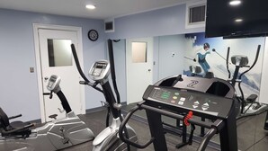 Uma academia com diversos equipamentos de exercícios, incluindo esteiras e elípticos, um espelho grande e uma televisão fixada na parede.