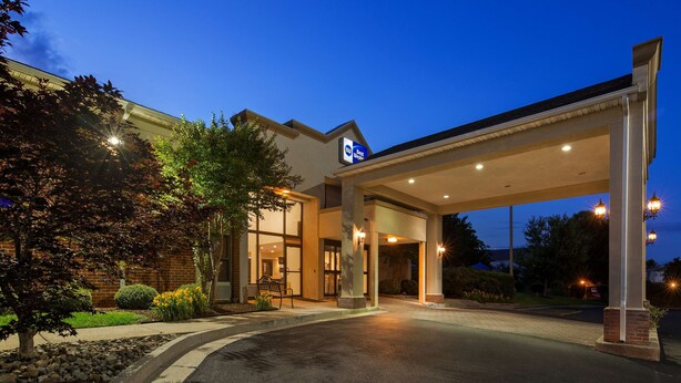 Entrada de um hotel Best Western ao entardecer, com área de estacionamento coberta, espaço para sentar ao ar livre e paisagismo bem cuidado.
