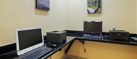 Uma secretária com um computador, um laptop e uma impressora. Existem duas molduras com quadros na parede acima da secretária.