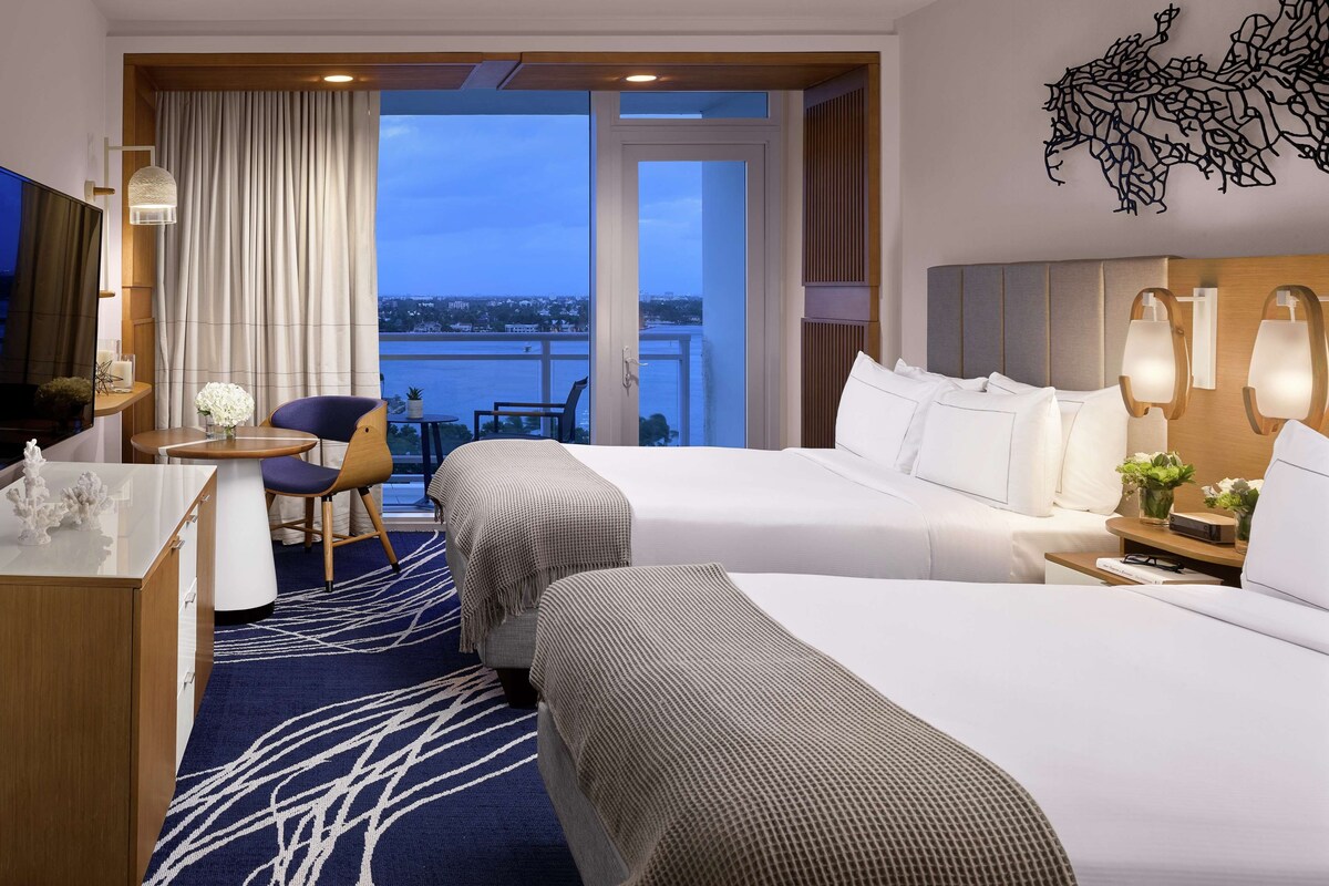 Quarto de hotel com duas camas, uma escrivaninha, uma cadeira, uma pequena mesa com um vaso de flores e vista para a cidade através de grandes janelas.