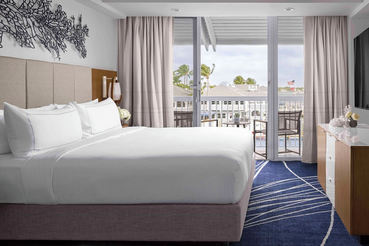 Quarto de hotel com uma cama grande, uma mesa, uma cadeira, vista para o oceano e varanda com guarda-corpo.