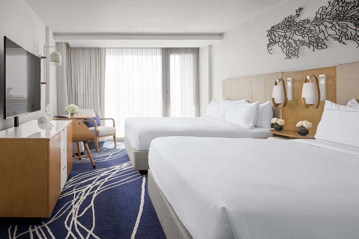 Quarto de hotel com duas camas, uma escrivaninha, televisão e uma janela grande com cortinas.