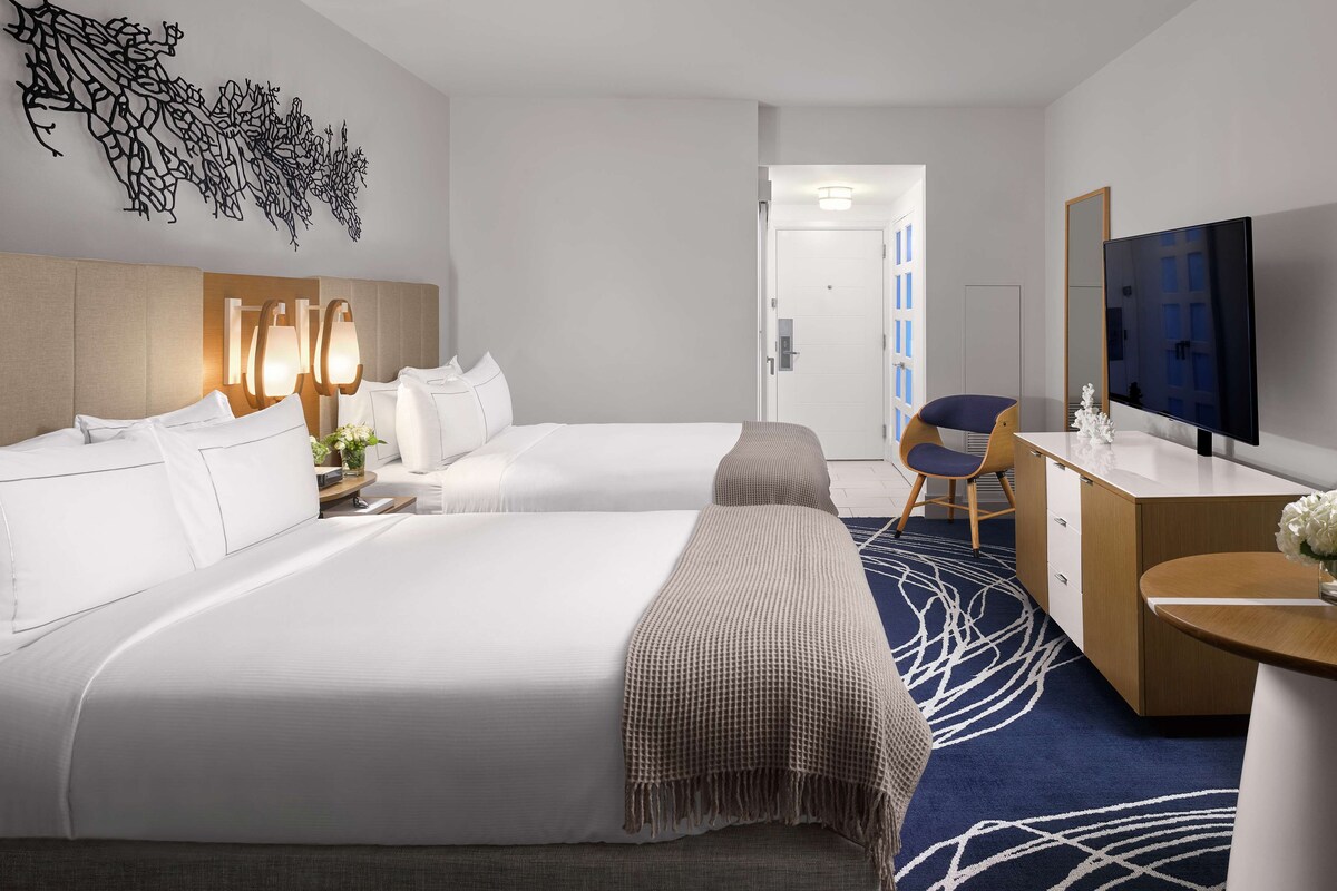 Quarto de hotel com duas camas, uma mesa de trabalho com televisão, uma cadeira e um objeto decorativo na parede.