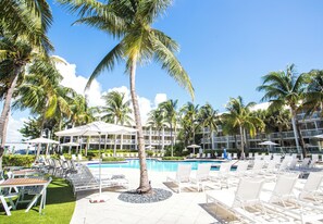 Uma área de piscina com espreguiçadeiras e guarda-sóis, cercada por palmeiras e um edifício com sacadas.