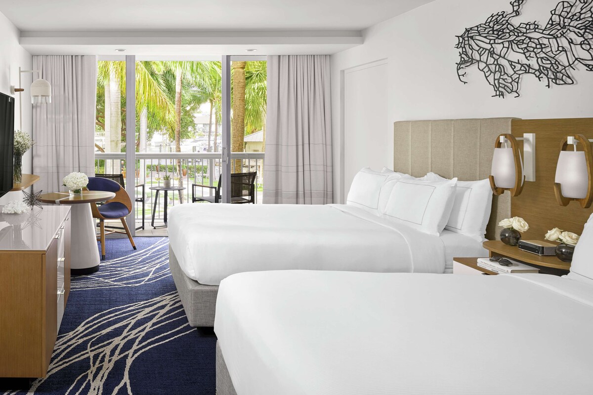 Quarto de hotel com duas camas, uma escrivaninha com cadeira, televisão e varanda com vista para palmeiras.