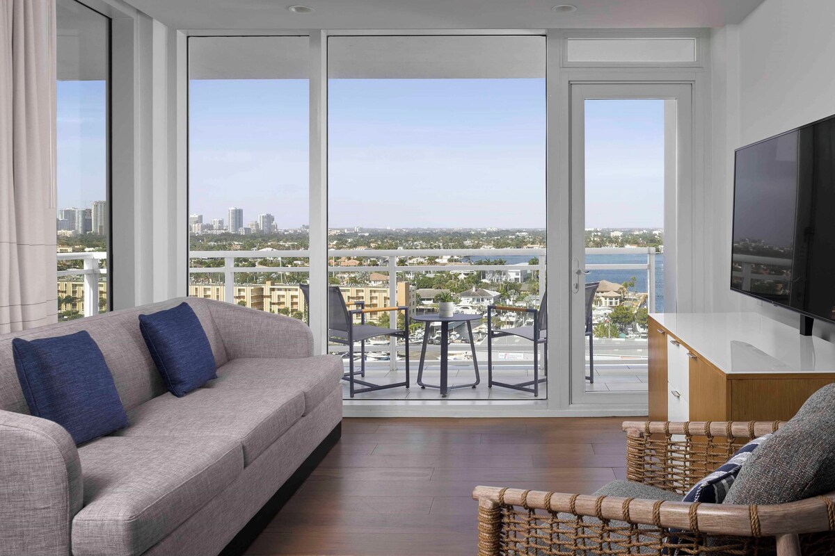 Uma sala de estar moderna com um sofá cinzento, uma televisão e vista para o horizonte urbano e para a água.
