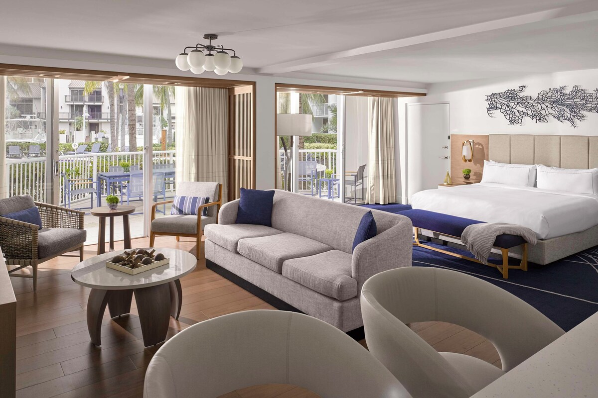 Quarto de hotel com cama, sofá, poltrona, mesinha e varanda com mobiliário.