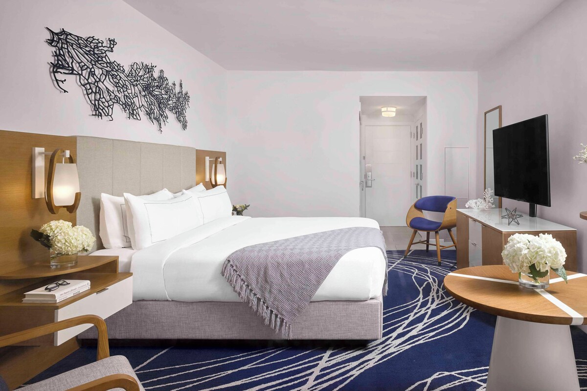 Quarto de hotel moderno com uma cama grande, uma escrivaninha com televisão de tela plana, uma cadeira e um painel decorativo na parede.
