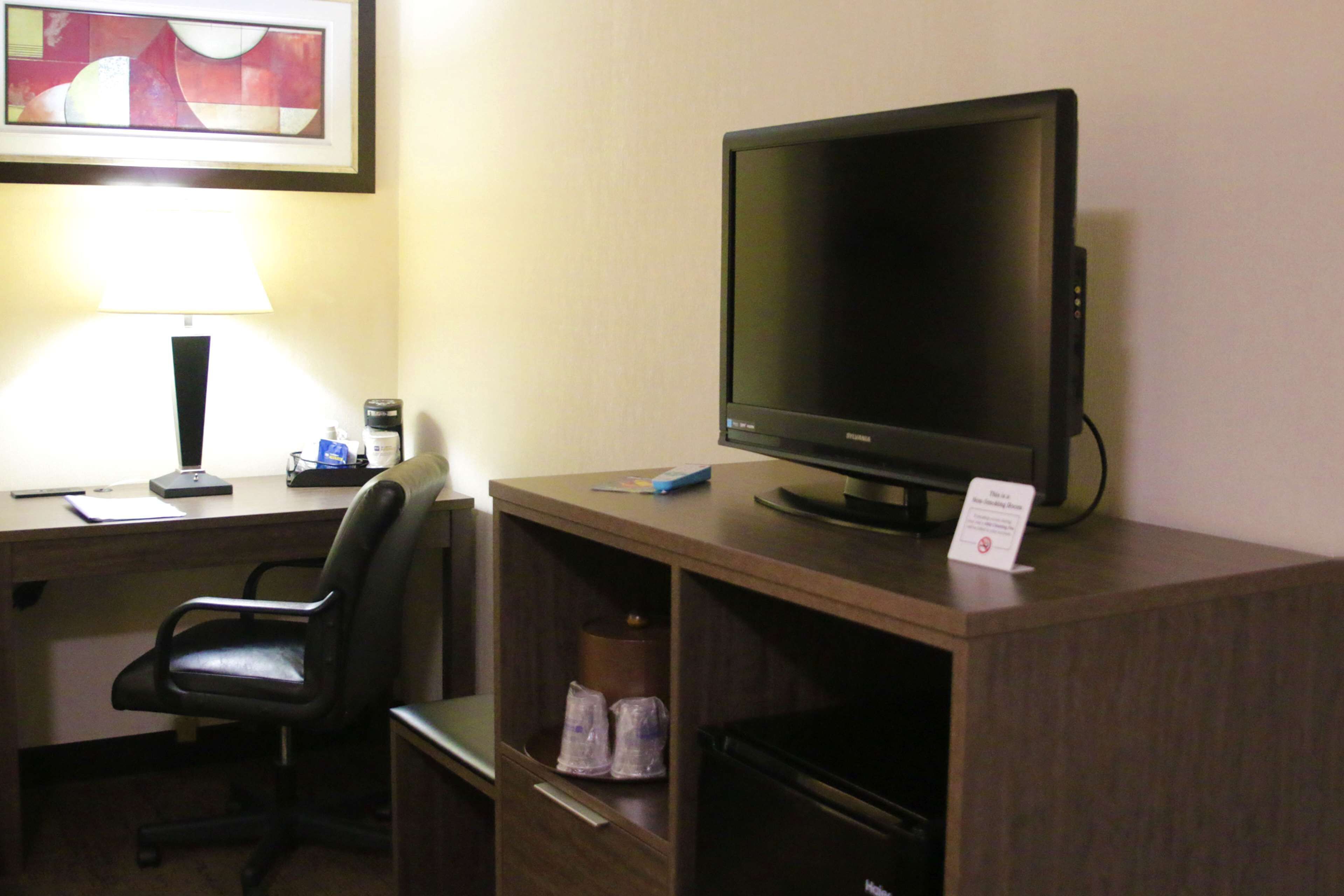 Quarto de hotel com escrivaninha, computador, cadeira, refrigerador e um quadro na parede.