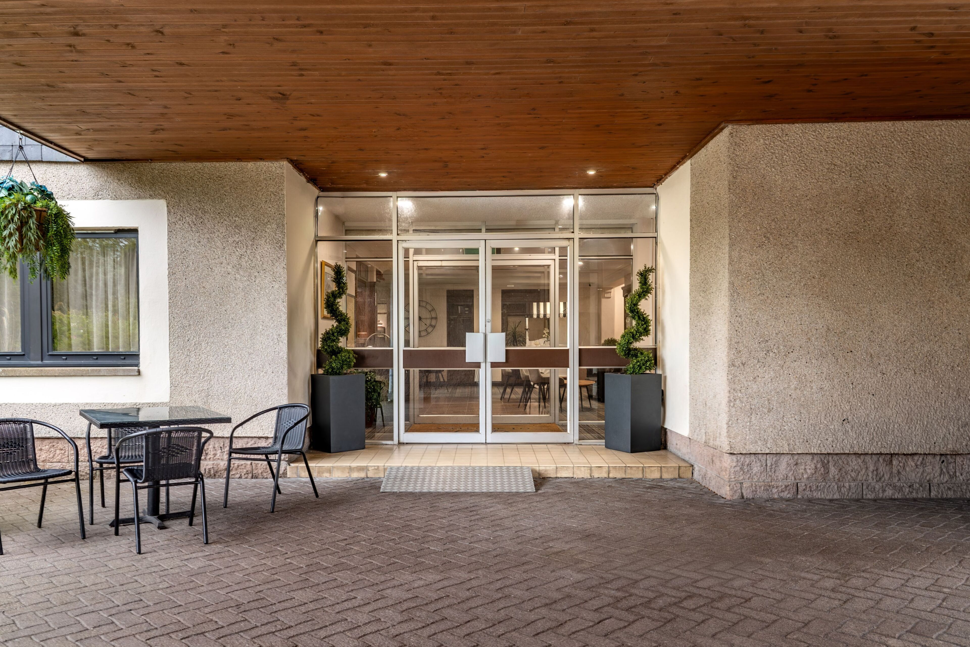 Entrada de hotel com porta de vidro, área de estar com cadeiras e uma mesa, e plantas em vasos.