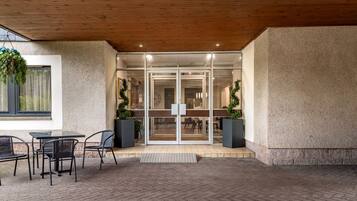 Entrada de hotel com porta de vidro, área de estar com cadeiras e uma mesa, e plantas em vasos.