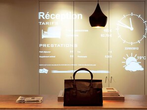 Uma área de recepção com um painel digital exibindo tarifas e serviços, um relógio e uma bolsa sobre uma mesa.