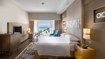 Quarto de hotel com cama, escrivaninha, cadeira, televisão e vista para o mar.
