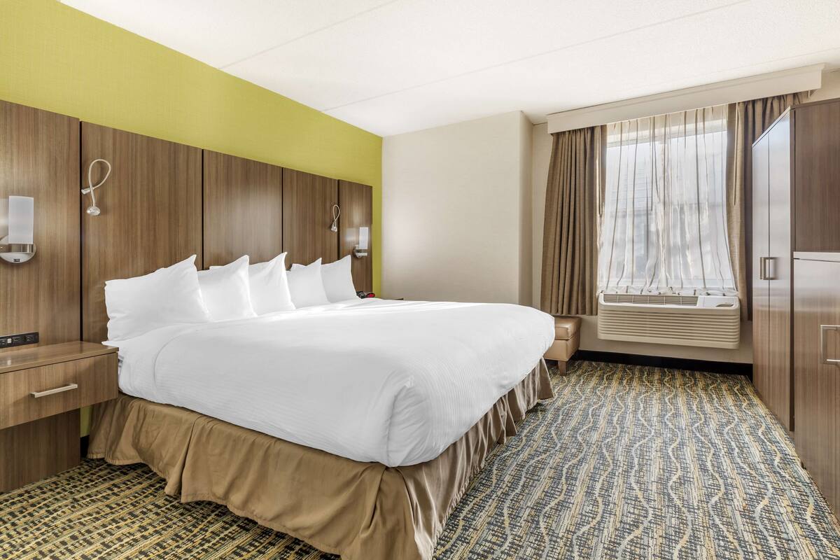Quarto de hotel com cama grande, cabeceira de madeira, mesa de cabeceira, cortinas e um tapete estampado.