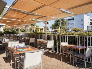 Área de terraço ao ar livre com mesas e cadeiras, protegida por grandes guarda-sóis, e cercada por uma grade de metal.