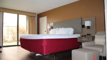 Quarto de hotel com cama grande, uma cadeira, escrivaninha e porta de vidro deslizante para uma varanda.