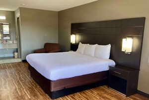 Quarto de hotel moderno com uma cama grande, cabeceira escura, duas luminárias de cabeceira, piso de madeira e um pequeno espaço de estar.