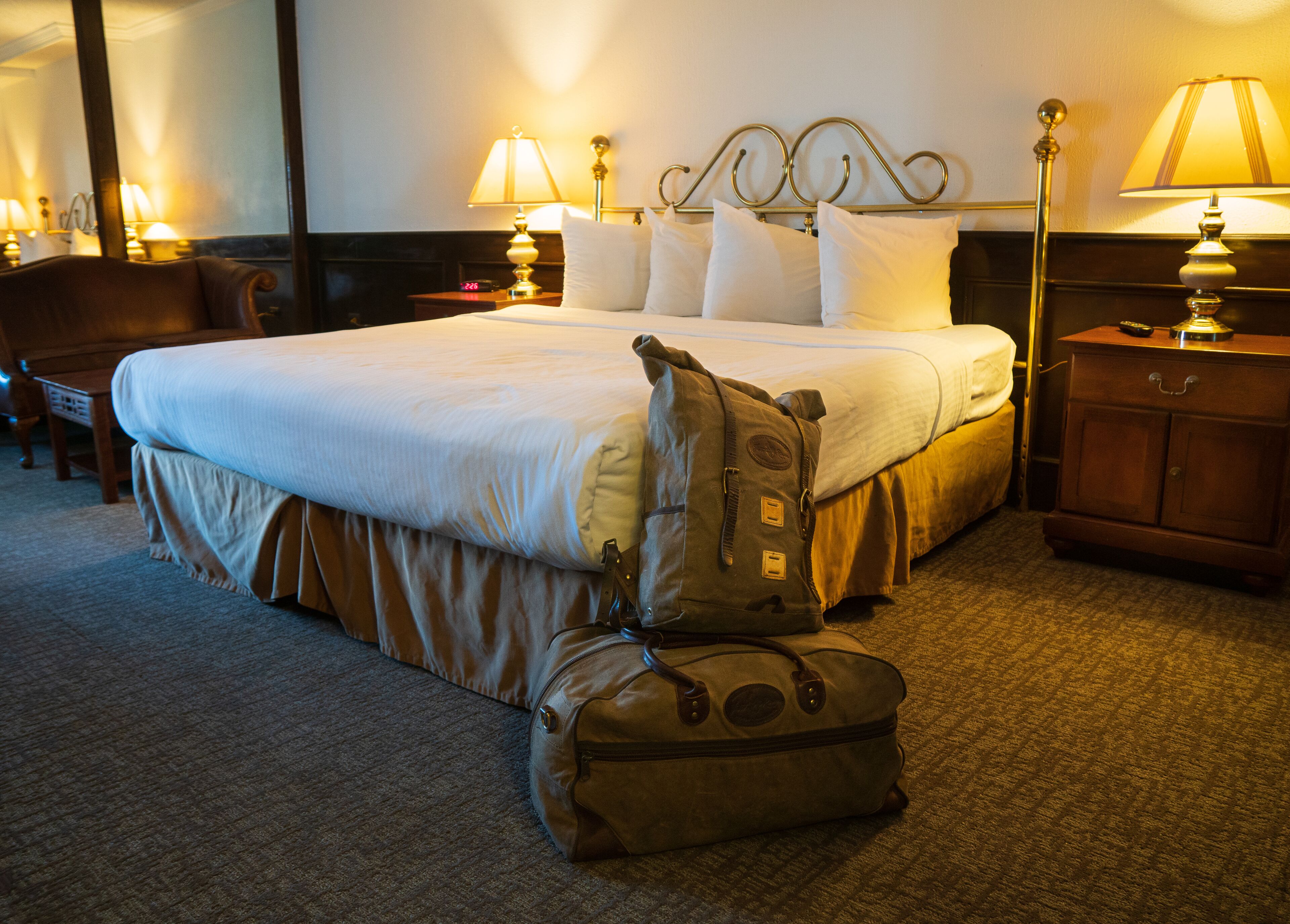 Quarto de hotel com uma cama grande, duas mesas de cabeceira com abajures, uma cômoda de madeira, uma cadeira de madeira e uma mala no chão.