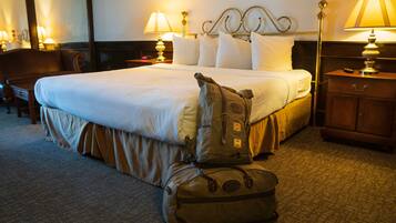 Quarto de hotel com uma cama grande, duas mesas de cabeceira com abajures, uma cômoda de madeira, uma cadeira de madeira e uma mala no chão.