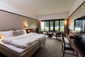 Quarto de hotel com uma cama grande, uma escrivaninha, uma cadeira e vista para uma paisagem verde através das janelas.