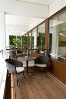 Varanda com cadeiras de vime e uma mesa, janelas amplas com vista para árvores e um deck de madeira.
