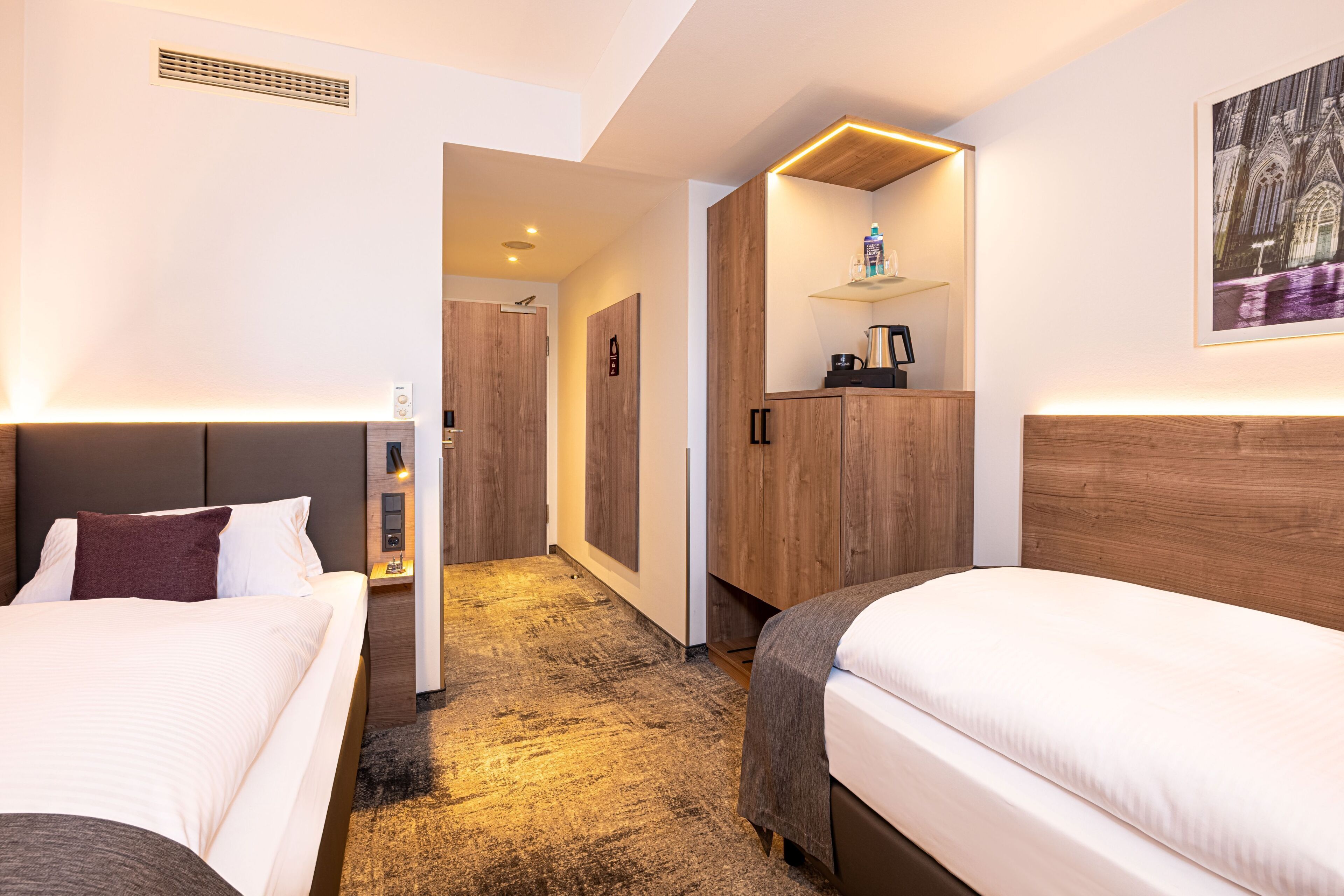 Quarto de hotel com duas camas, um guarda-roupa de madeira, uma pequena secretária com uma lâmpada e um quadro na parede.