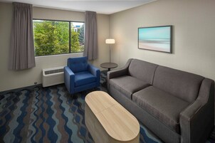 Quarto de hotel com uma janela grande, uma poltrona azul, um sofá cinzento, uma mesa redonda pequena e uma mesinha de cabeceira de madeira.