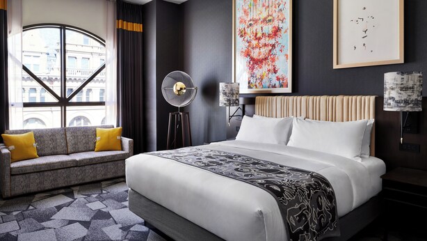 Quarto de hotel moderno com uma cama grande, um sofá com almofadas amarelas, um ventilador e obras de arte nas paredes.