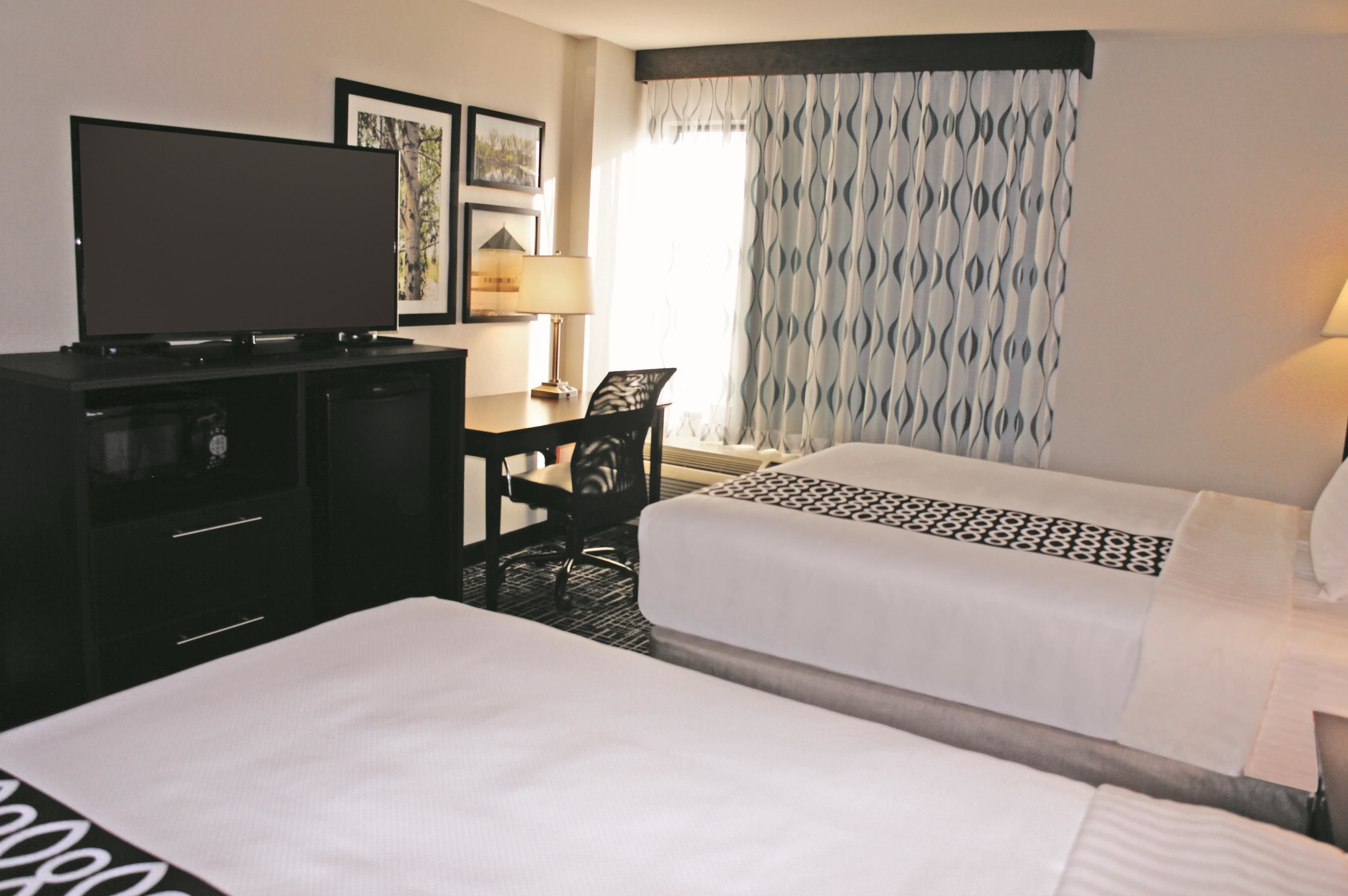 Quarto de hotel com duas camas, uma escrivaninha com cadeira, televisão e janela com cortinas estampadas.